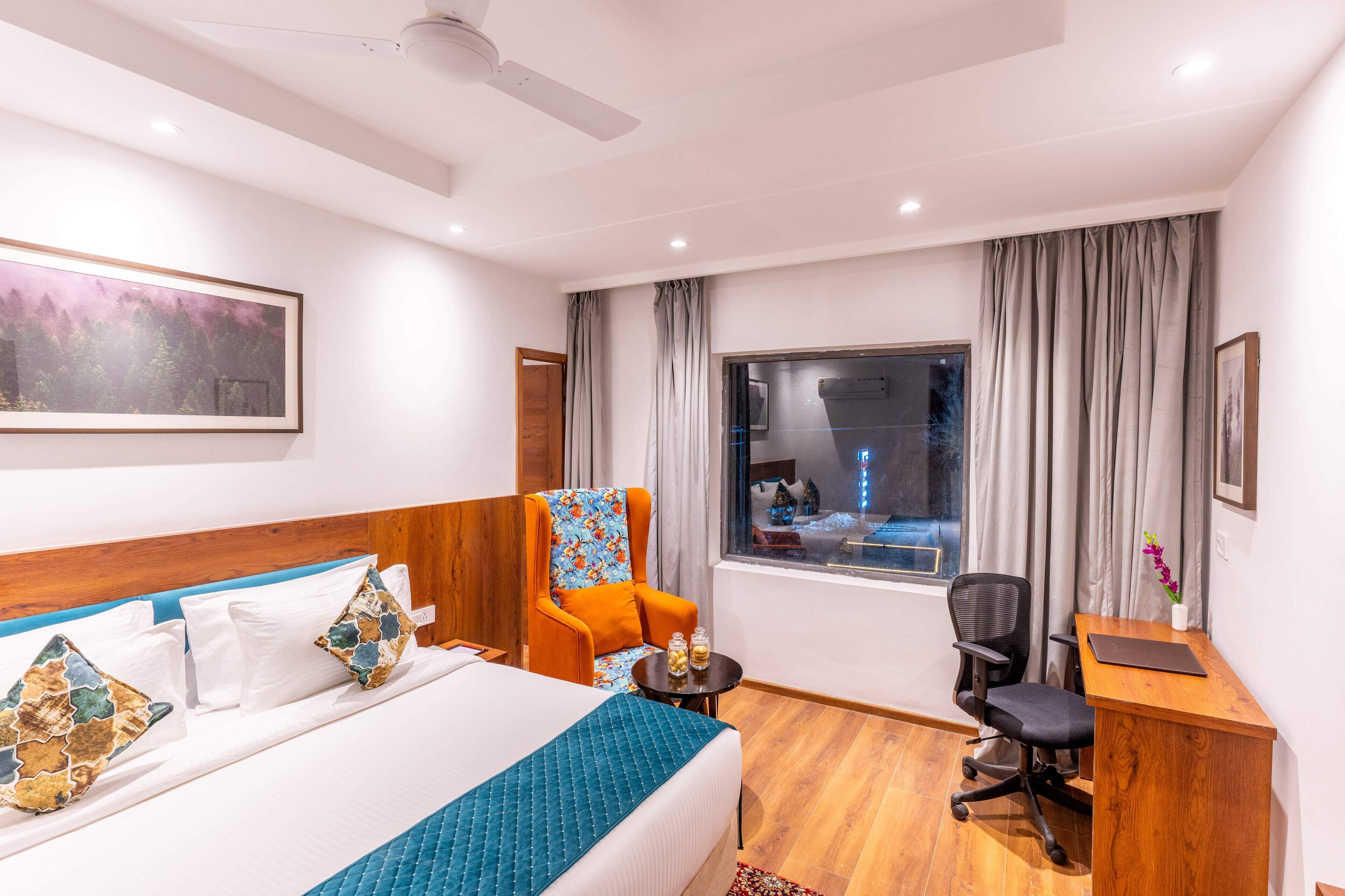 Quarto de hotel com cama, escrivaninha, cadeira, janela com cortinas e ventilador de teto.