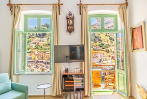 Quarto com uma televisão grande, um armário de madeira e vista para uma cidade colorida através de janelas com moldura verde.