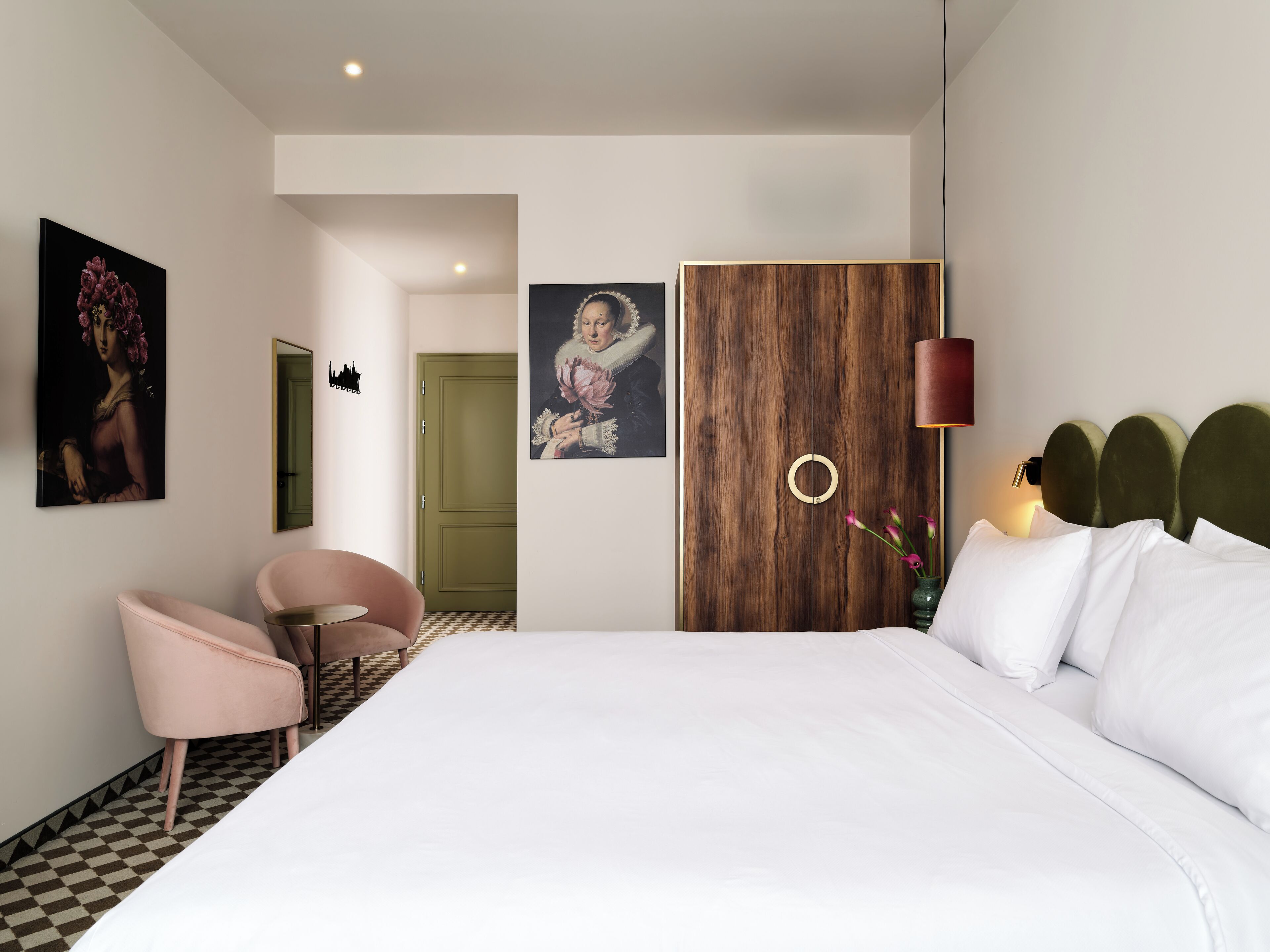 Quarto de hotel moderno com uma cama grande, cabeceira de madeira, uma poltrona rosa e duas pinturas penduradas na parede.
