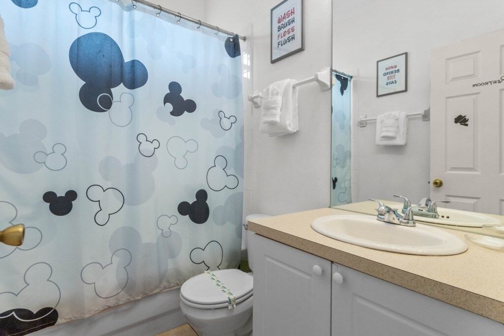 Um banheiro com cortina de chuveiro do Mickey Mouse, vaso sanitário, pia e espelho.