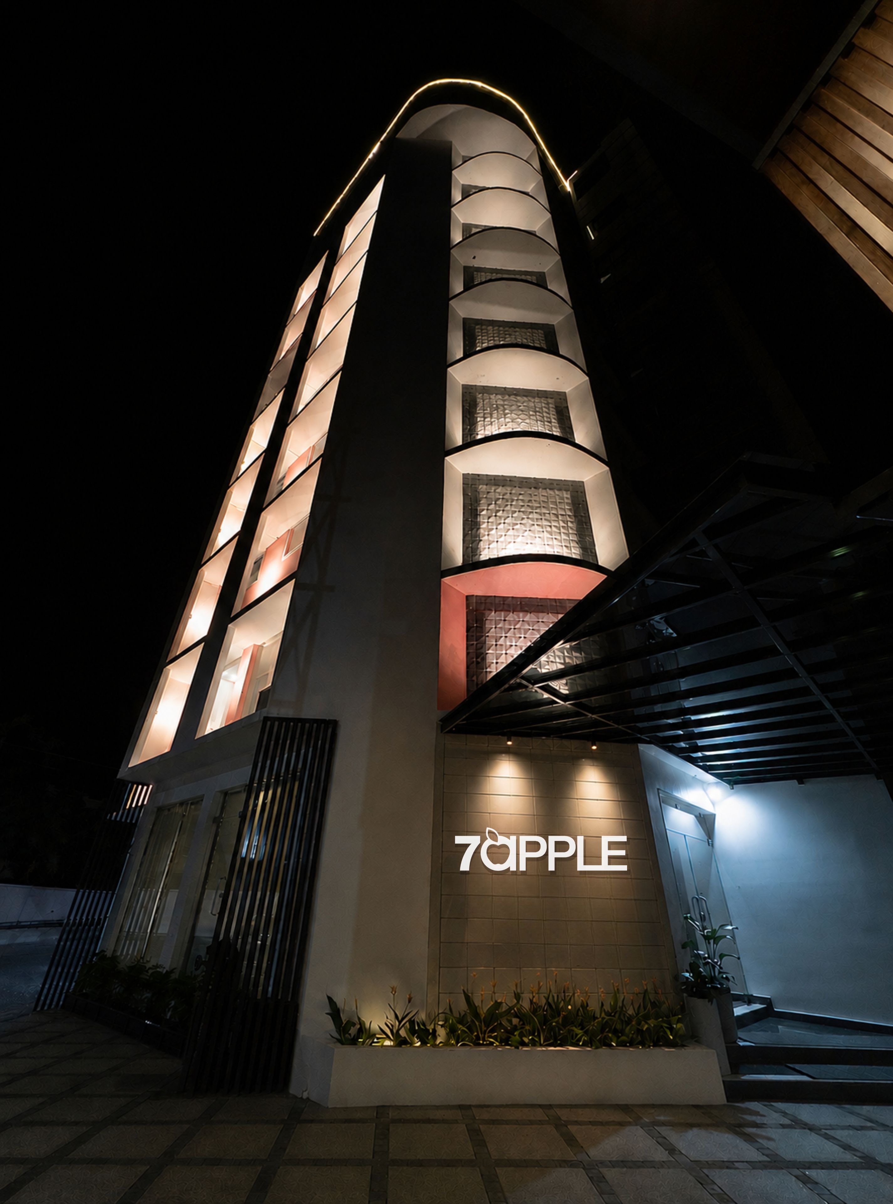 Um edifício moderno com fachada curva, janelas iluminadas e uma placa com a inscrição '7APPLE'.