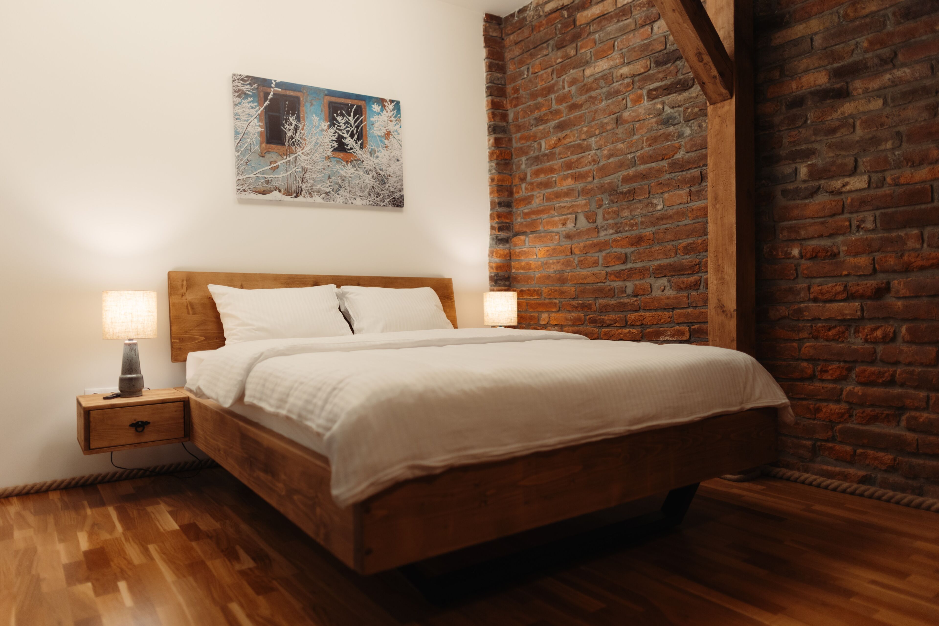 Quarto com cama de madeira, duas luminárias de cabeceira, mesa de apoio, parede de tijolos e um quadro na parede.