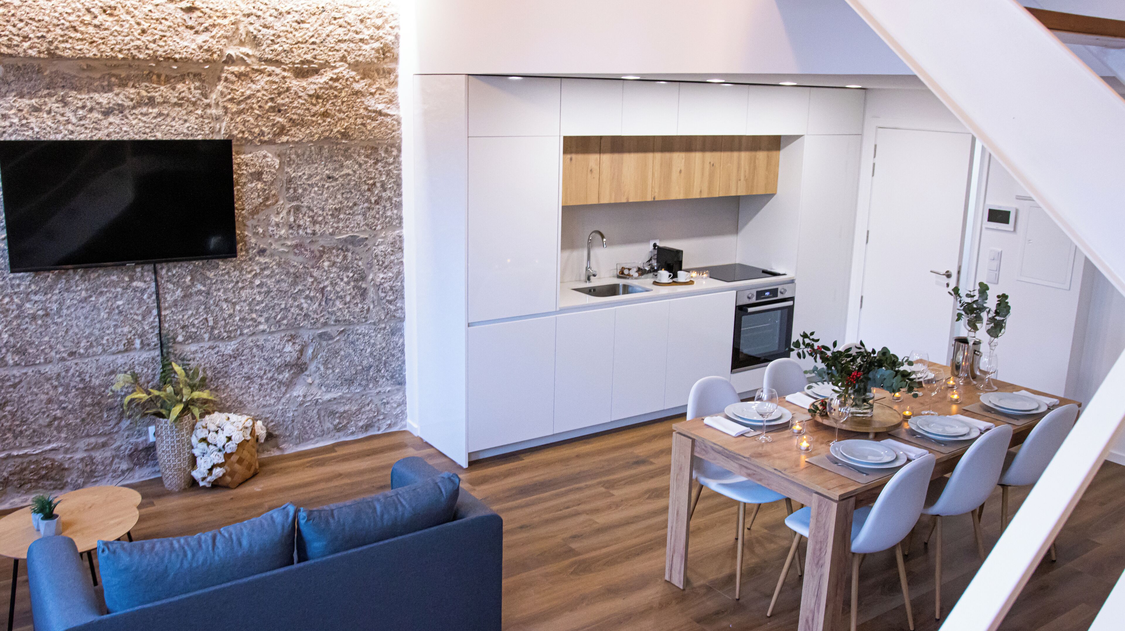 Uma cozinha moderna com armários brancos, bancada de madeira e forno embutido. Há uma área de jantar com mesa de madeira posta para quatro, cadeiras azuis e um pequeno vaso com planta.