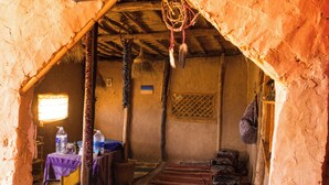 Um quarto rústico com telhado de palha, uma mesa com garrafas de água, um tapete estampado e uma cama com almofadas estampadas.