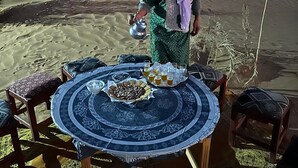 Cena de deserto com uma pessoa despejando água em um recipiente metálico, uma mesa com bandeja de comida e bebidas, e toalha de mesa estampada.