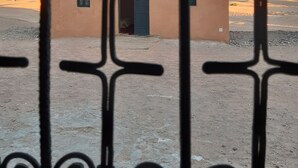 Um pequeno edifício com porta e janela, visto através de um portão de metal com detalhes decorativos.