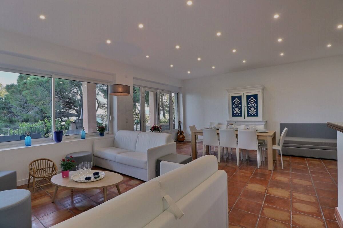 Ampla sala de estar com sofá, mesa de jantar e cadeiras. Grandes janelas oferecem iluminação natural e vista para o exterior.