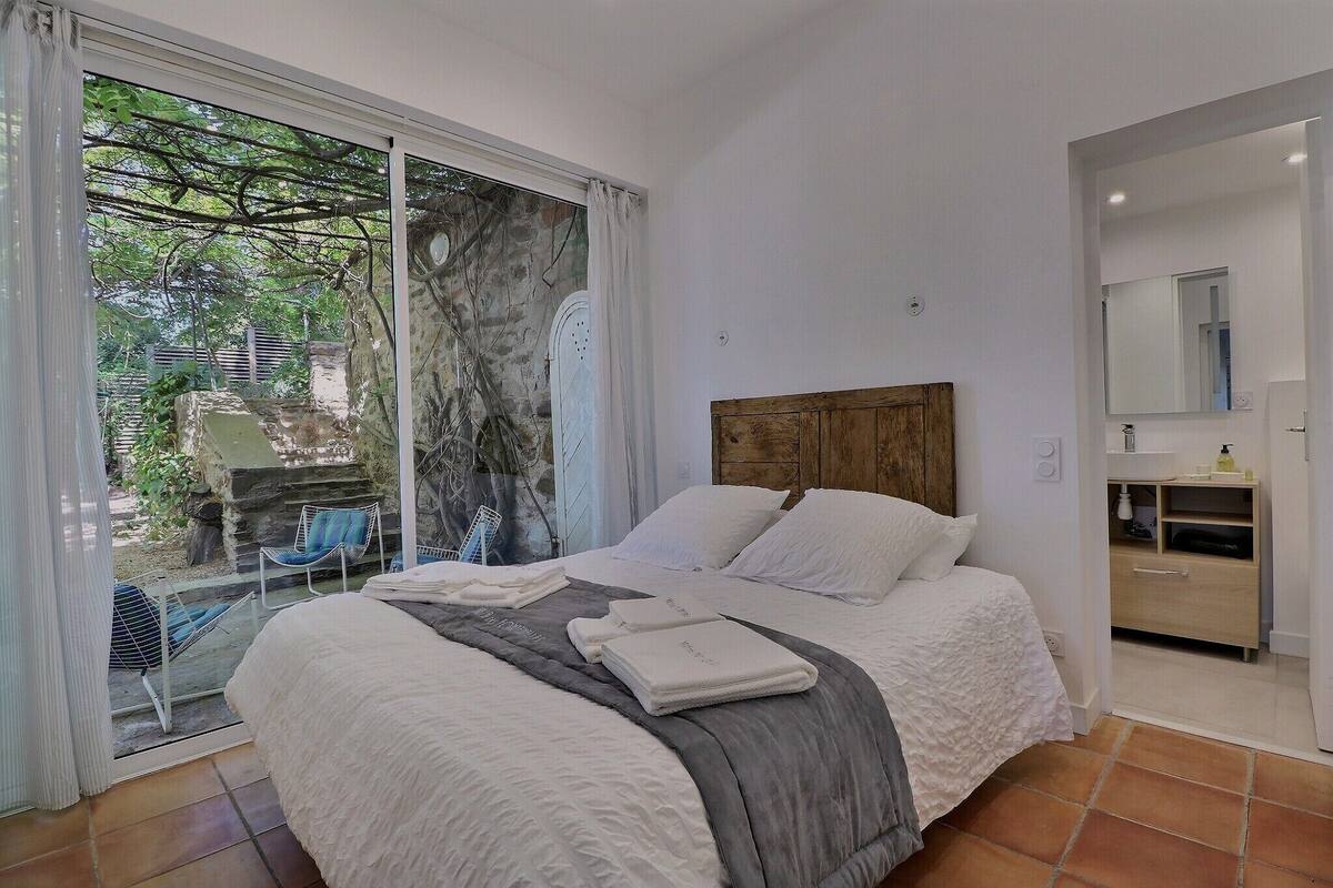 Quarto com cama grande, cabeceira de madeira, banheiro com espelho e pia, e vista para o verde pela janela.