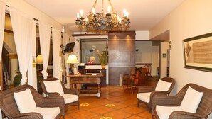 Um hall de hotel com cadeiras de vime, um candelabro, um balcão de recepção e piso de azulejos.