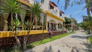 Uma casa de dois andares com fachada amarela e branca, uma varanda e um jardim com palmeiras e uma flor cor-de-rosa.