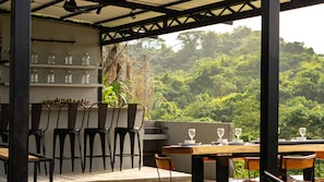 Uma área de refeições coberta ao ar livre, com cadeiras de metal e uma mesa posta para dois, com vista para uma floresta verde e exuberante.