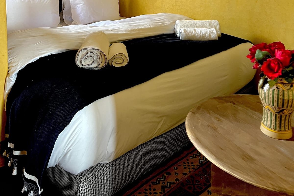 Quarto de hotel com cama, duas luminárias de cabeceira, um lustre decorativo, mesa de cabeceira com um vaso de flores e um tapete estampado.