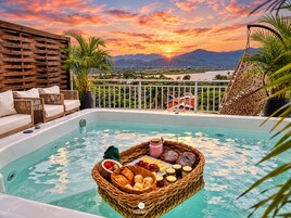 Piscina no terraço com vista para o pôr do sol, móveis de vime e uma cesta de frutas.