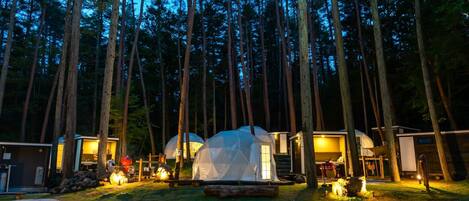 Uma área arborizada com várias cabanas em formato de cúpula, um caminho de cascalho e uma fogueira.