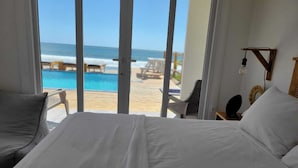 Quarto com uma cama grande, vista para o oceano e área de piscina visível pela janela.