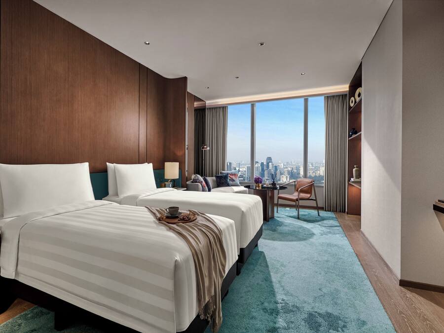 Quarto de hotel com duas camas, uma escrivaninha, uma cadeira, televisão e uma janela grande com vista para a cidade.