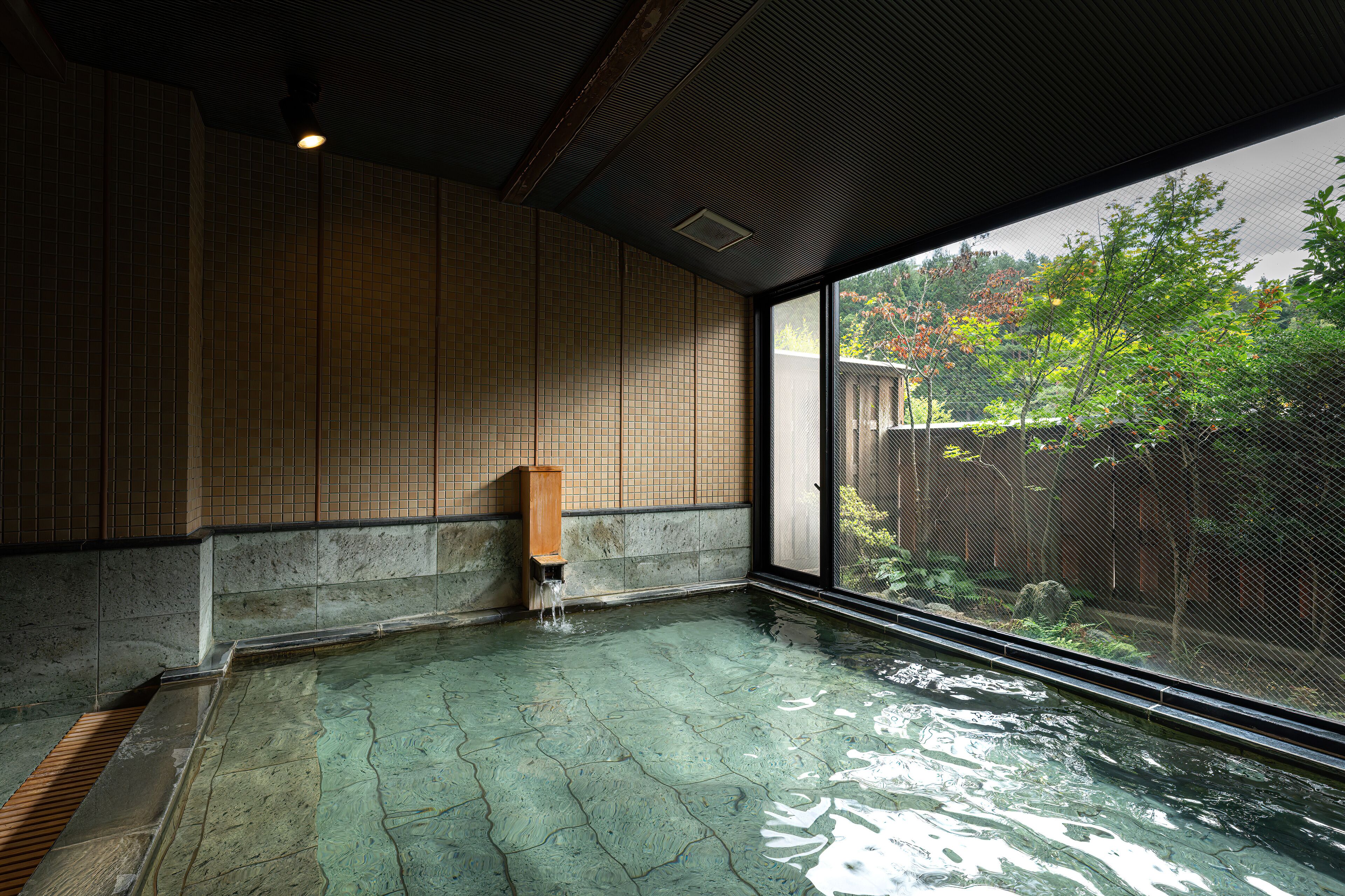 Uma banheira onsen japonesa tradicional, com parede e banco de madeira, e vista para um jardim por uma grande janela.