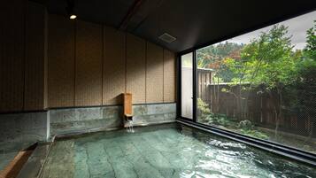 Uma banheira onsen japonesa tradicional, com parede e banco de madeira, e vista para um jardim por uma grande janela.