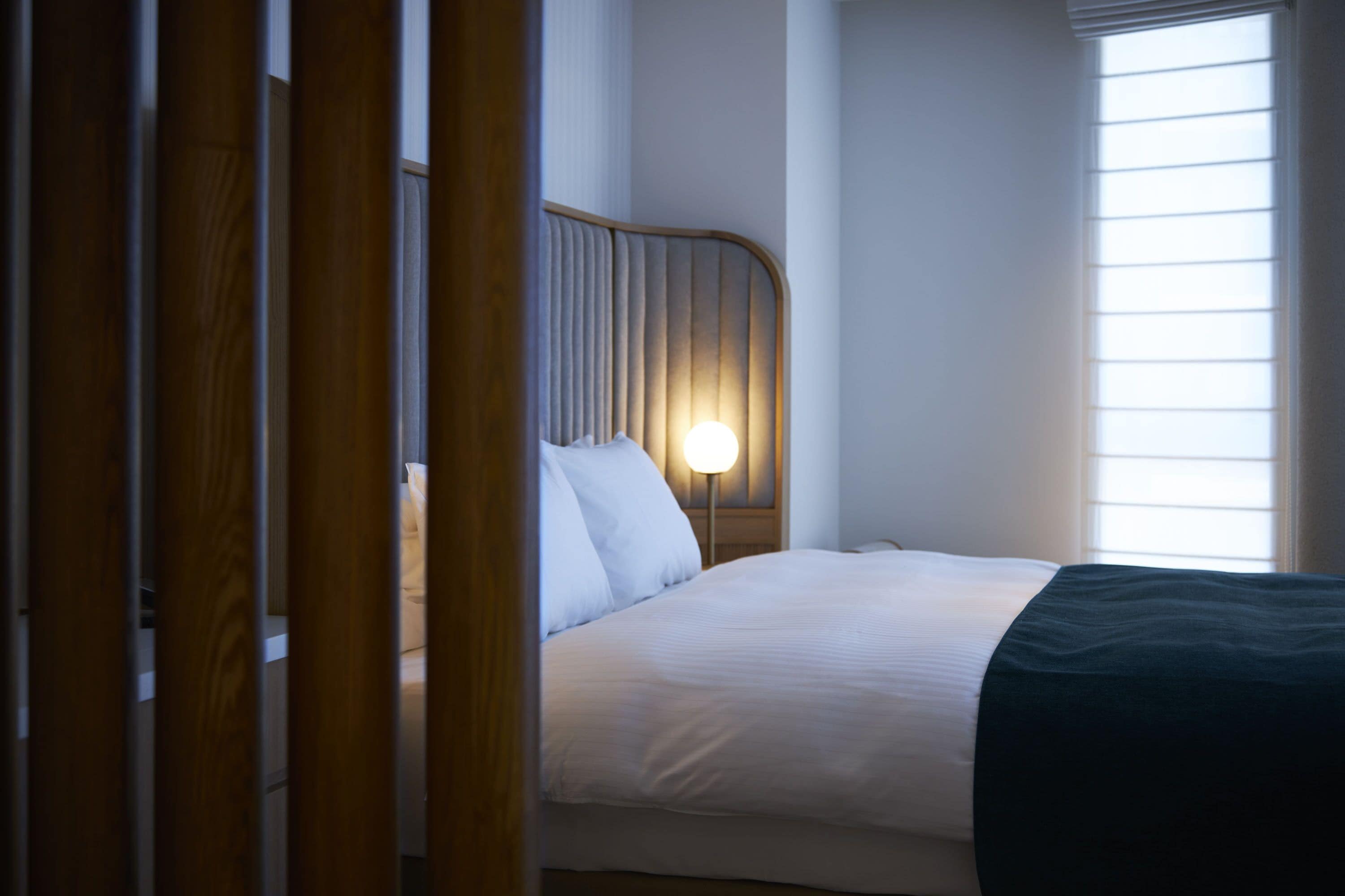 Cama bem arrumada com cabeceira de madeira, abajur e janela com persianas.