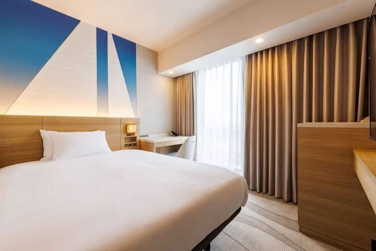 Quarto de hotel com uma cama grande, uma escrivaninha e uma cadeira, uma janela com cortinas, e uma parede com um desenho geométrico.