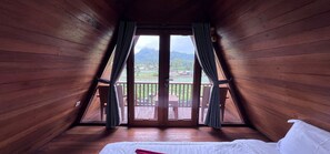 Quarto com cama, paredes de madeira e vista para uma paisagem montanhosa por uma porta de correr.
