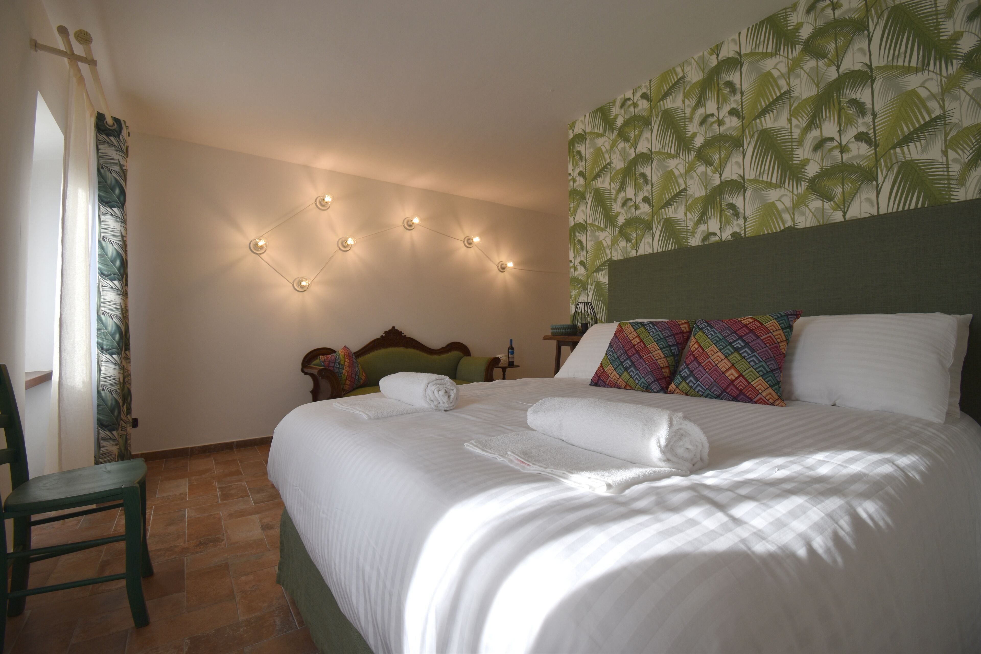 Uma cama bem arrumada com roupa de cama branca, cabeceira verde e papel de parede estampado ao fundo.