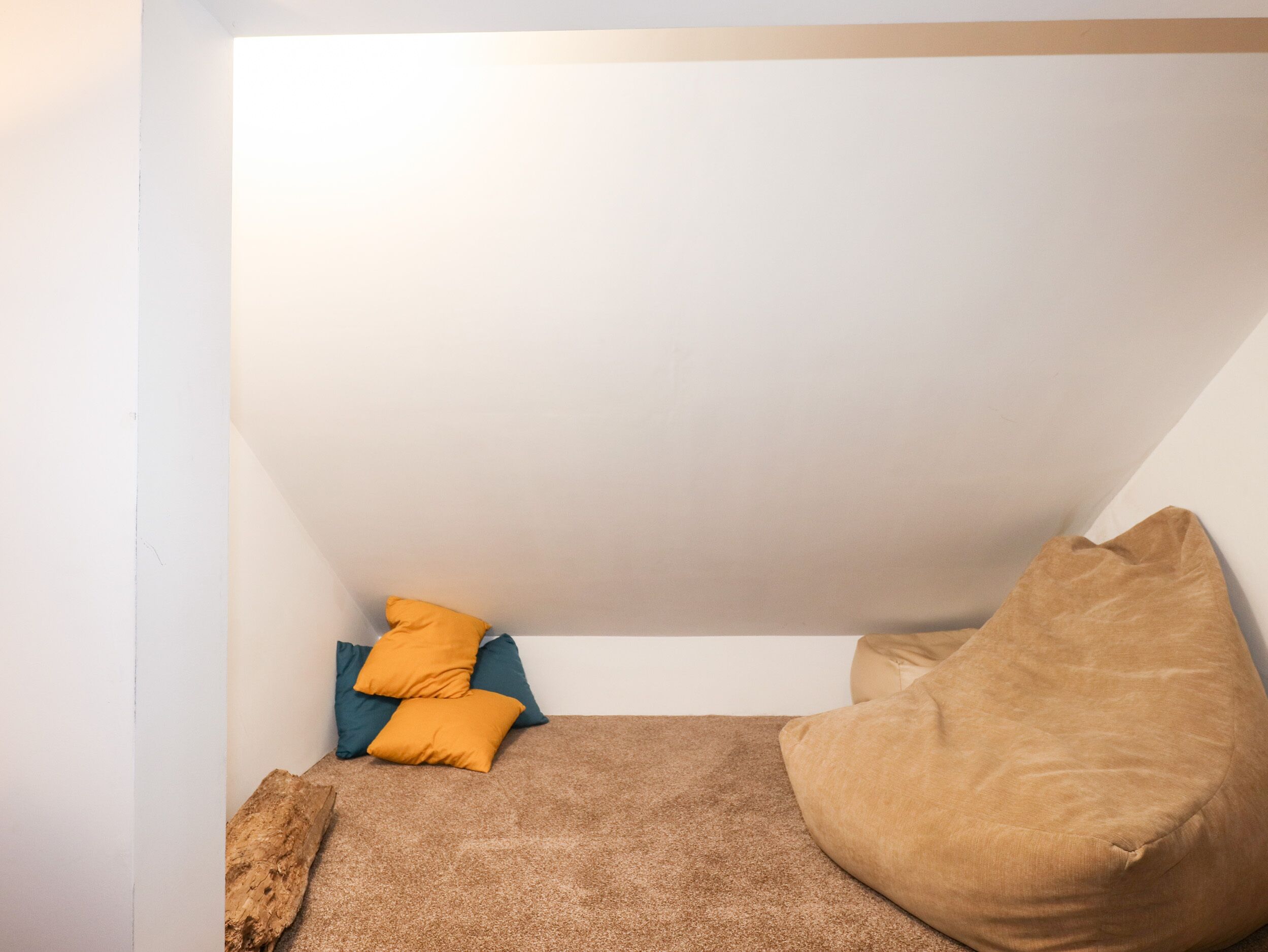 Um quarto pequeno e acolhedor com teto inclinado, carpete marrom e dois travesseiros (um laranja, um azul) sobre uma plataforma baixa.