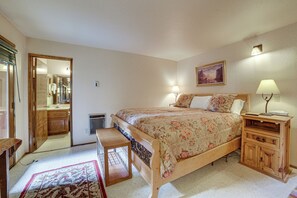 Quarto com cama de madeira, mesas de cabeceira, um criado-mudo, um espelho e uma porta para um banheiro.