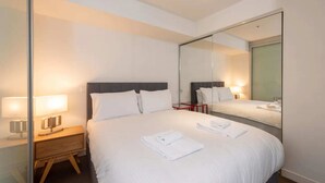 Quarto de hotel com uma cama grande, uma mesa de cabeceira com abajur, um espelho e toalhas dobradas com cuidado.