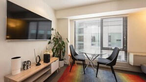 Uma sala de estar moderna com uma televisão de tela plana na parede, um console de madeira, duas cadeiras pretas, uma mesa de centro de vidro e uma janela grande com persianas.