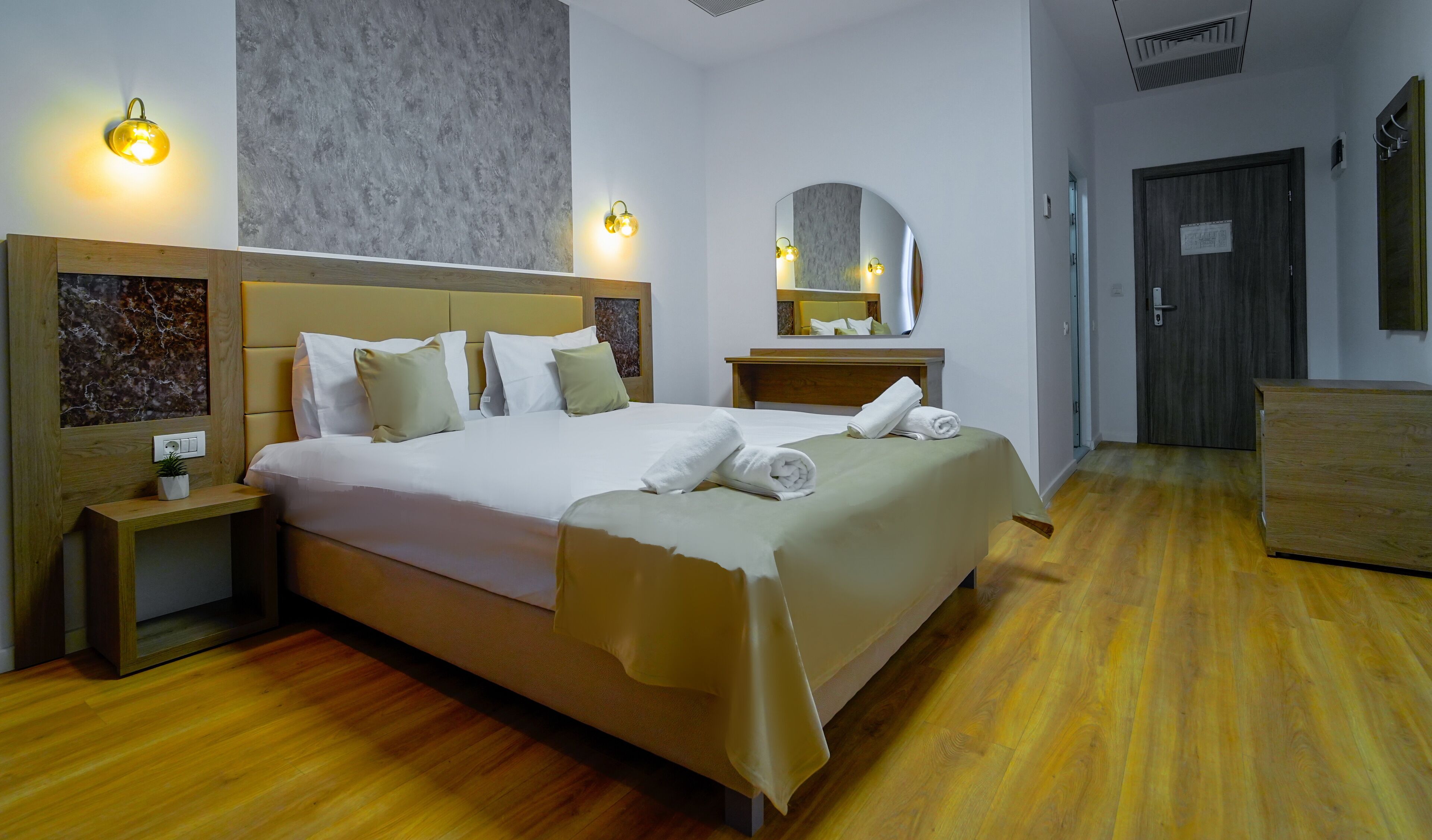 Quarto de hotel com uma cama grande, mesas de cabeceira, cômoda de madeira, espelho e arandelas.