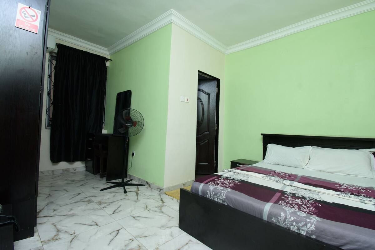 Um quarto com cama, escrivaninha, cadeira, ventilador e porta.