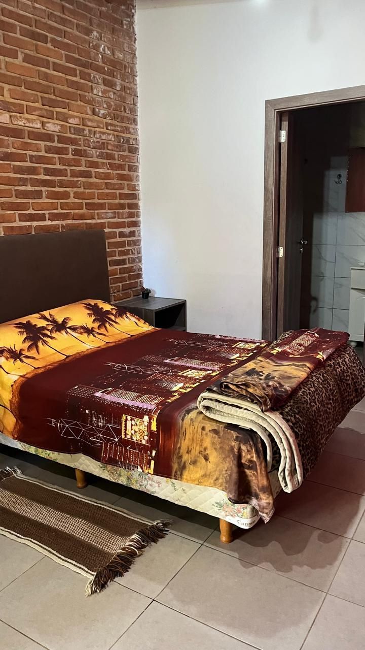 Uma cama com colcha estampada, uma mesa de cabeceira, uma parede de tijolos e uma porta para um banheiro.
