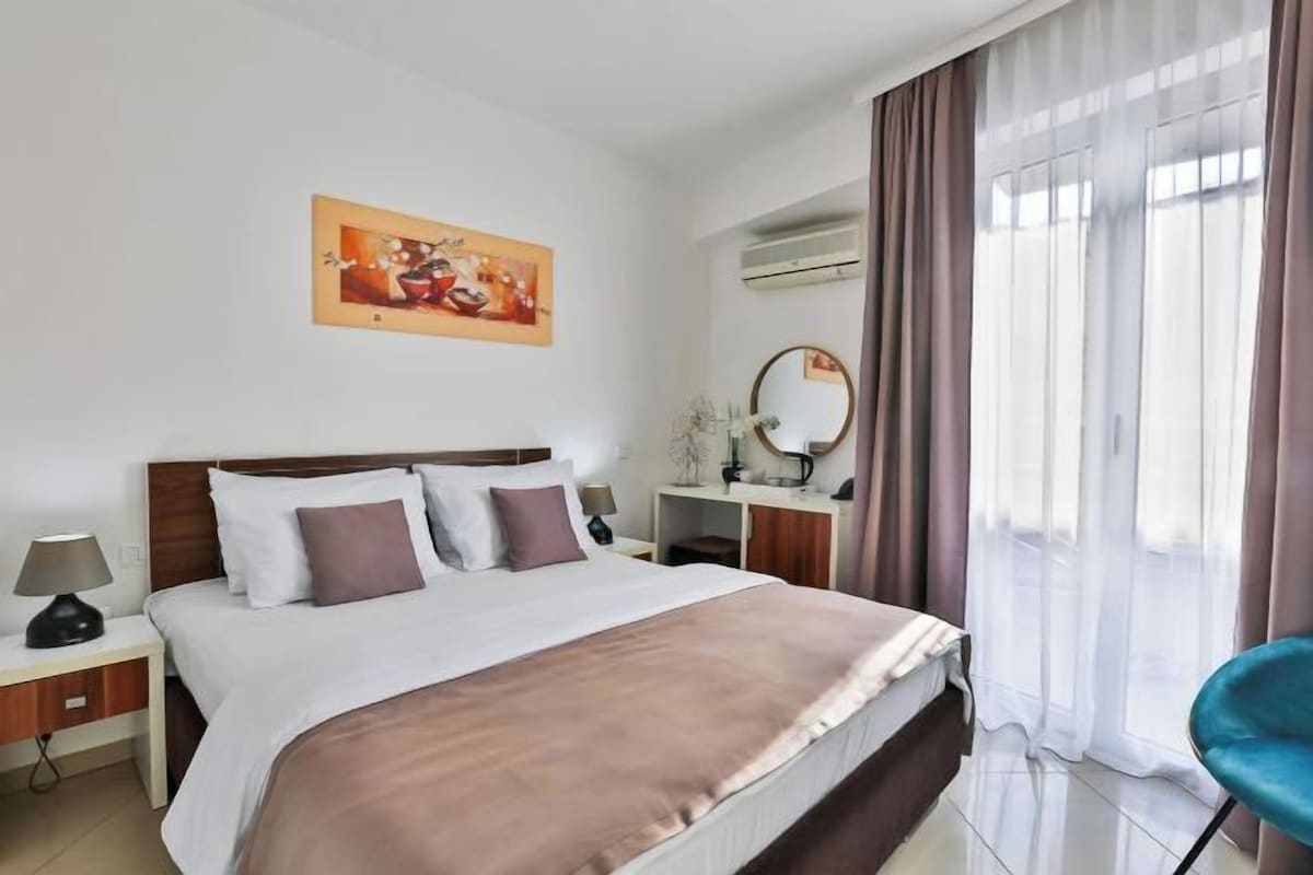 Quarto de hotel com cama, mesas de cabeceira, escrivaninha, cadeira e janela com cortinas.