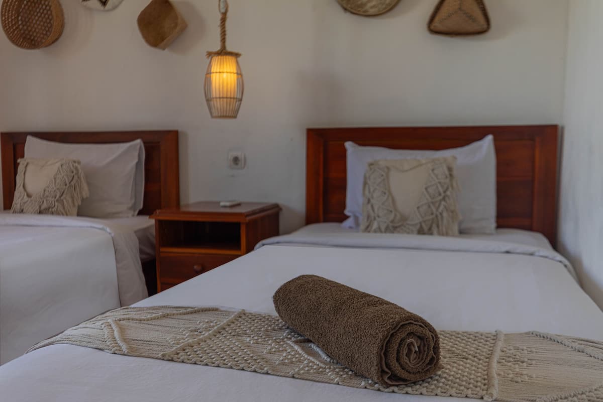 Quarto de hotel com duas camas de solteiro, uma mesinha de cabeceira de madeira, um tapete tecido e decoração de parede incluindo cestos de vime e um lustre.