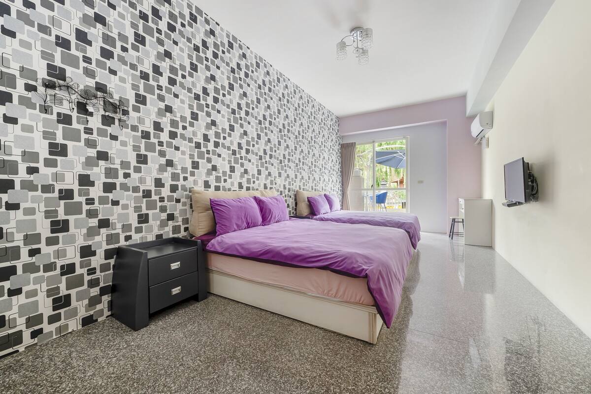 Quarto moderno com parede estampada, cama com roupa de cama roxa, mesa de cabeceira preta e televisão na parede.