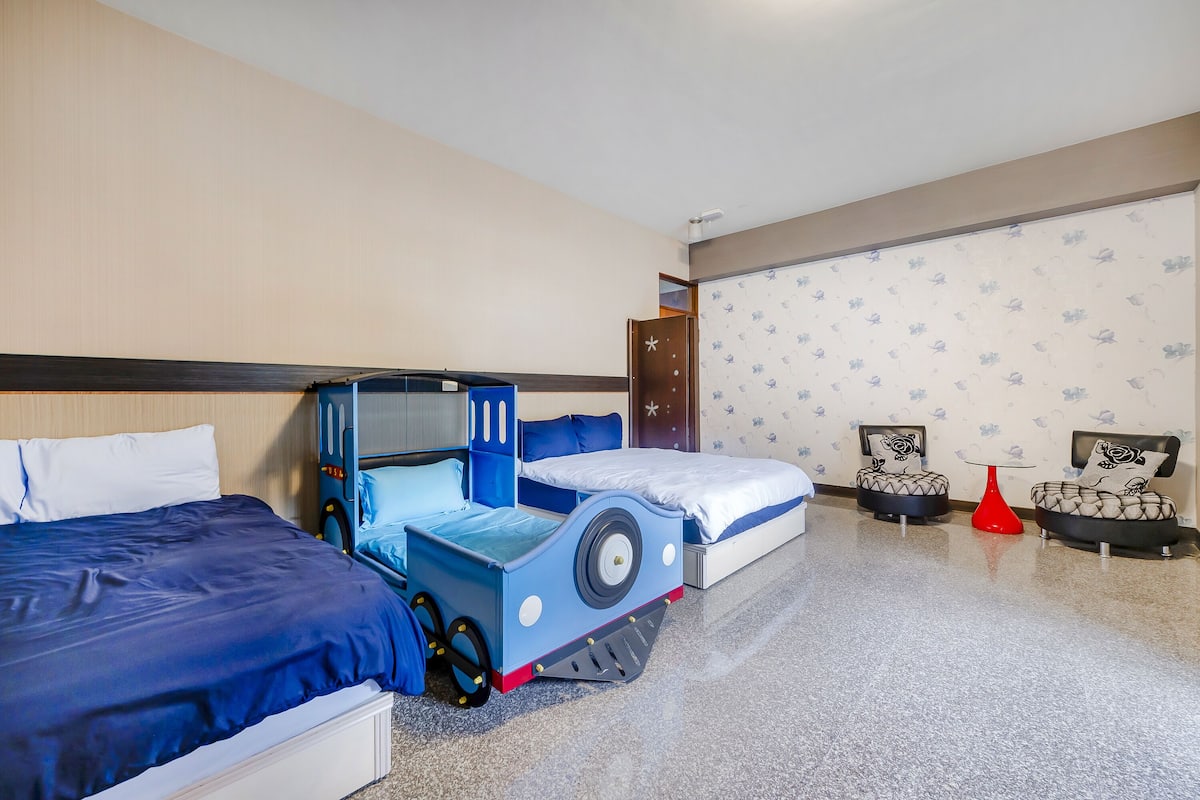 Quarto de hotel com duas camas, incluindo uma cama com tema de trem, uma mesa vermelha e uma parede estampada.