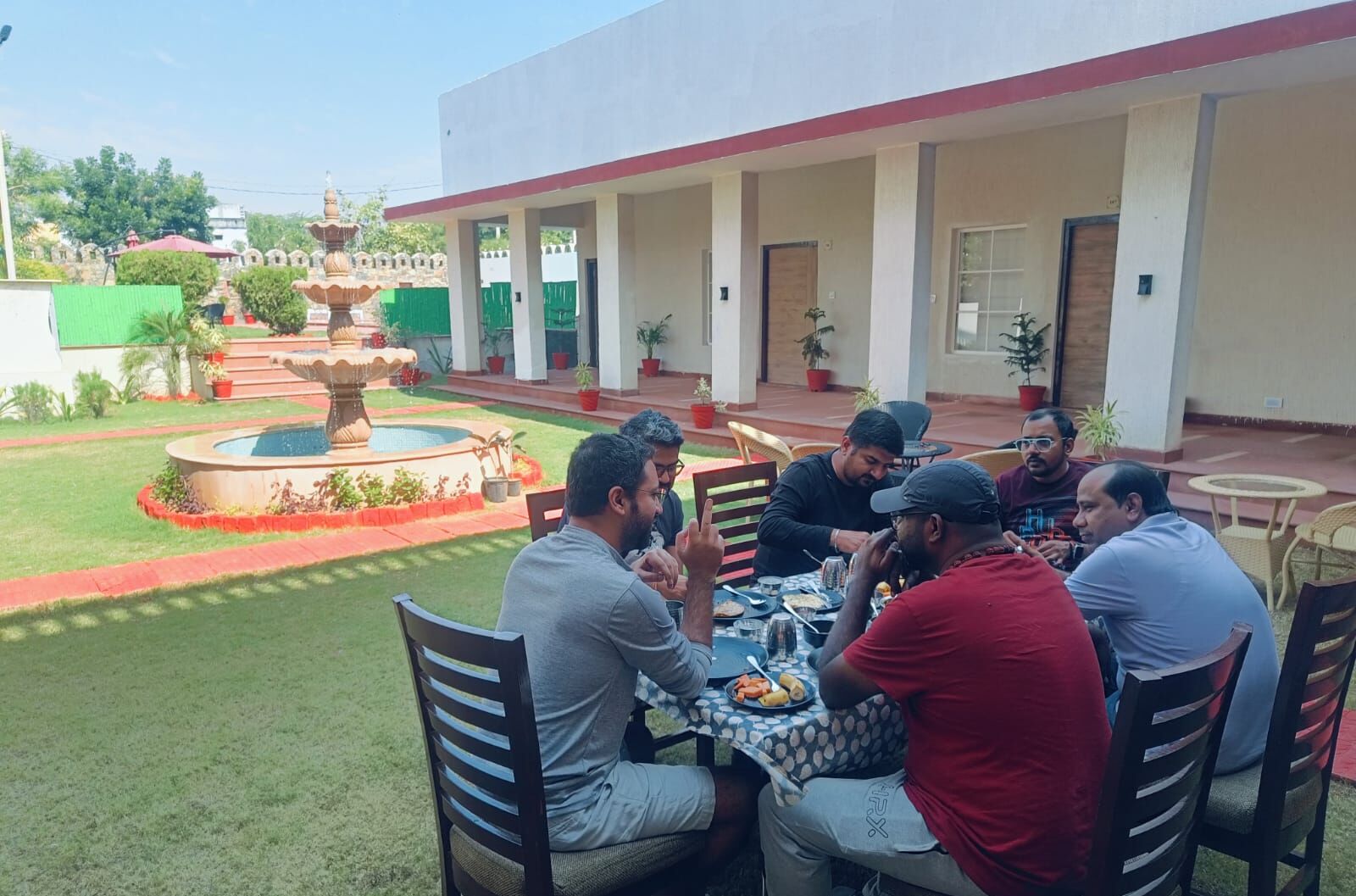 Um grupo de pessoas jantando ao ar livre em um restaurante, com uma fonte e um jardim ao fundo.