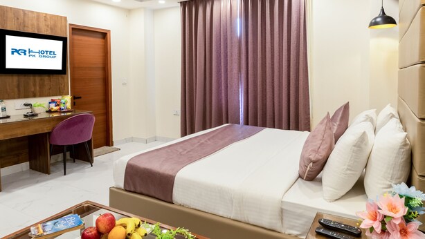 Quarto de hotel com cama, escrivaninha, cadeira, mesa de centro de vidro com frutas e uma planta, televisão e ventoinha de teto.