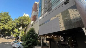 Um edifício moderno com telhado plano e grandes janelas, próximo a uma rua com carros estacionados e uma pequena árvore.