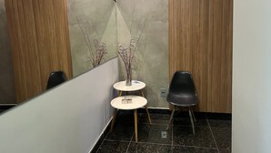Espaço de espera pequeno e moderno, com duas mesas redondas, uma cadeira preta e um espelho que reflete o ambiente.