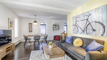 Uma sala de estar moderna com área de jantar, um sofá com almofadas, uma mesa de centro de madeira e uma grande obra de arte na parede retratando uma bicicleta.
