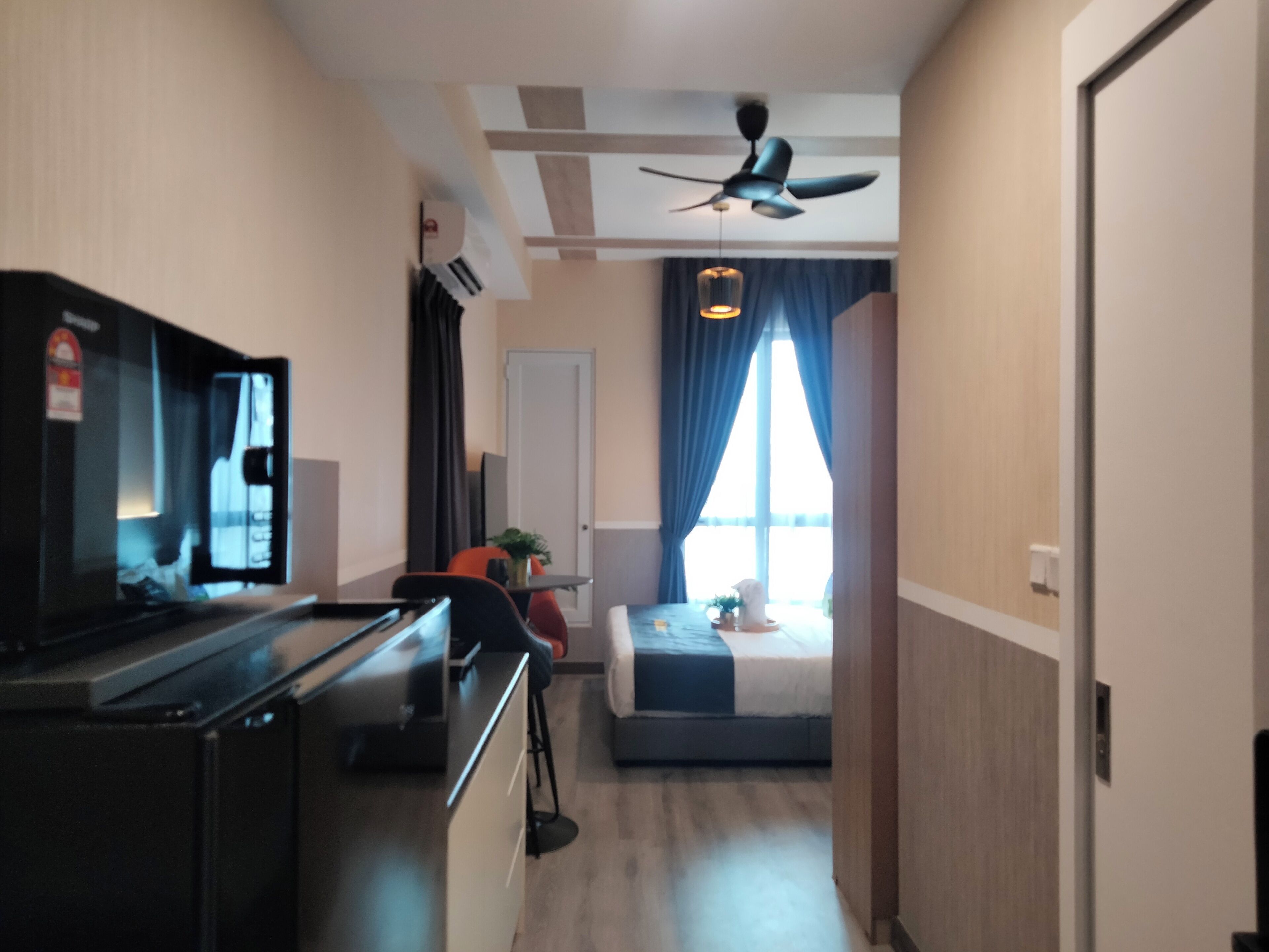 Quarto de hotel compacto com cozinha compacta, uma cama, ventoinha no teto e uma janela grande com cortinas.