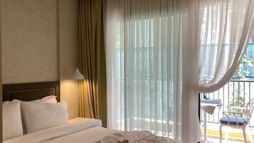 Quarto de hotel com uma cama grande, cabeceira, abajur, janela com cortinas leves e vista para a varanda externa.