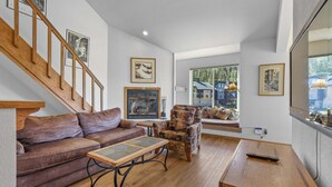 Uma sala de estar ampla com escada de madeira, lareira, sofá, poltrona estampada e uma grande janela com vista para o exterior.