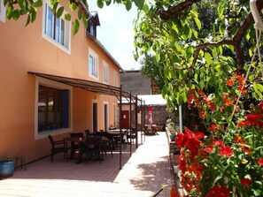 Uma área de pátio ao ar livre com mesas e cadeiras, um edifício com janelas e um jardim com flores vermelhas.