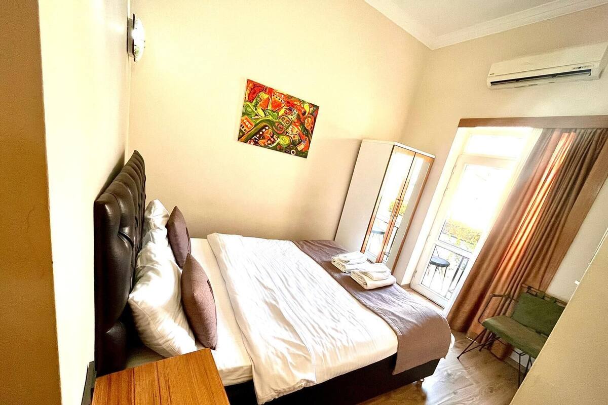 Quarto de hotel com uma cama, mesa de cabeceira de madeira, um espelho e uma janela com cortinas.