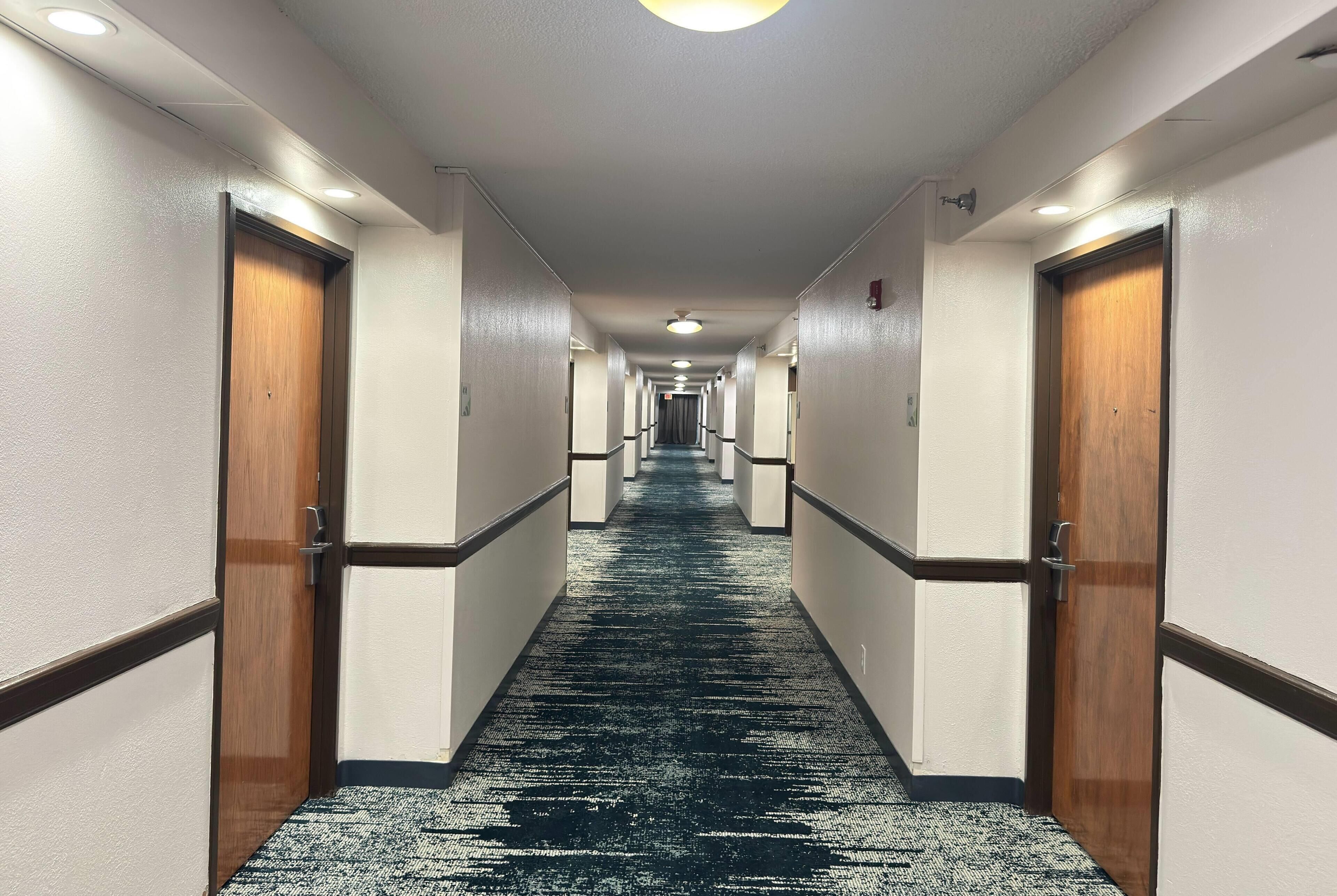 Corredor de hotel com portas em ambos os lados, piso coberto de carpete e iluminação embutida.
