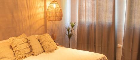 Quarto com uma cama grande, um banquinho, luminária pendente e cortinas.