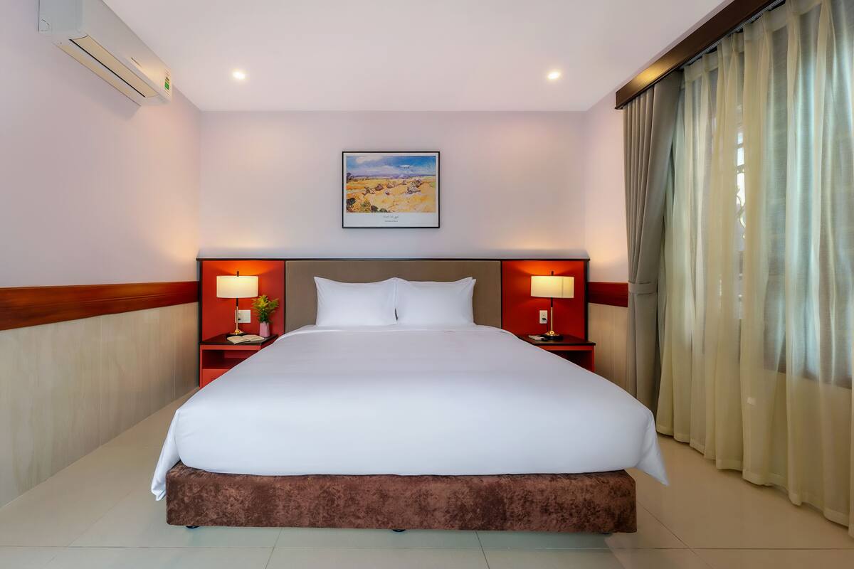 Quarto de hotel com uma cama grande, duas mesas de cabeceira com luminárias, ar condicionado fixado na parede, um quadro emoldurado e uma janela com cortinas.